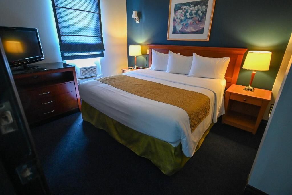 Quarto de hotel com uma cama grande, duas mesas de cabeceira com abajures, uma TV, uma janela com persianas e um quadro na parede.