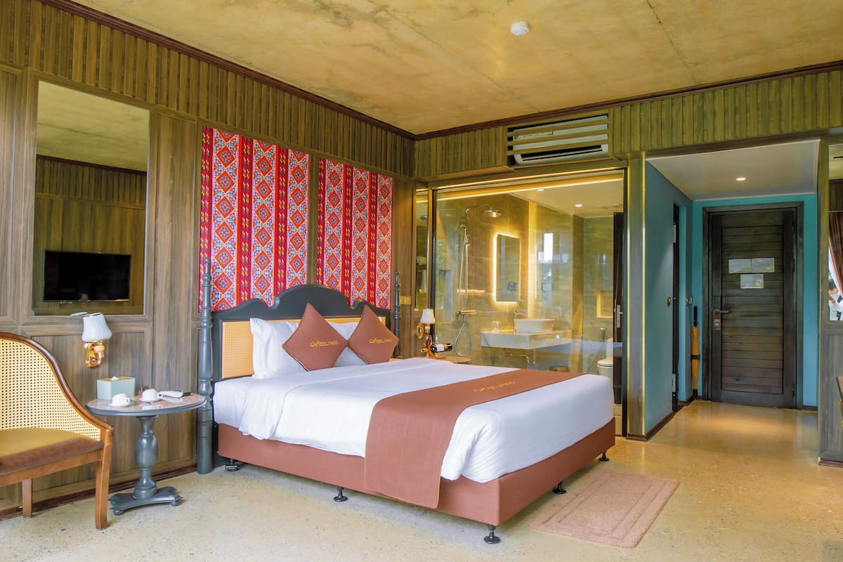 Quarto de hotel com uma cama grande, mesa de cabeceira com luminária, uma cadeira e uma televisão.