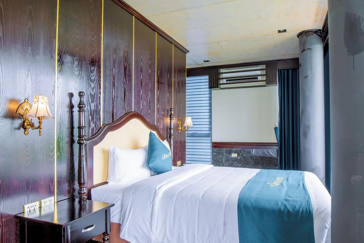 Quarto de hotel com uma cama grande, cabeceira de madeira, mesa de cabeceira e duas arandelas.