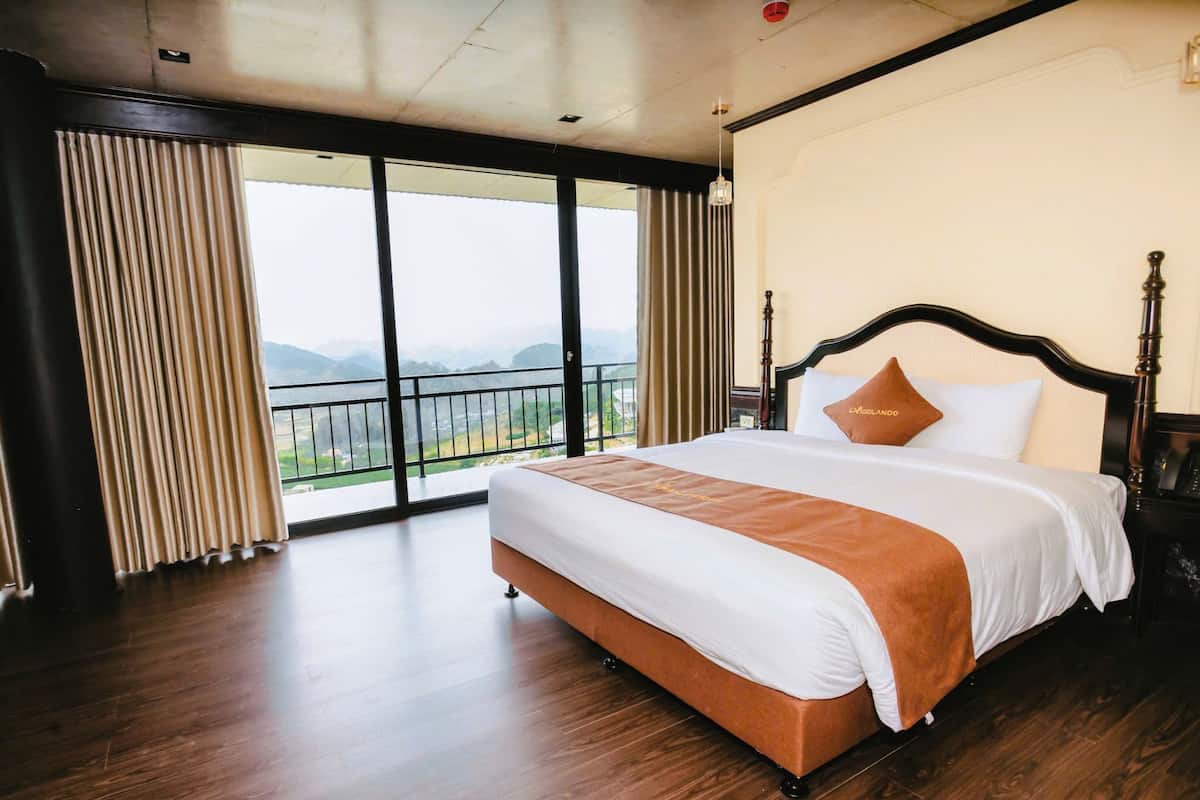 Uma cama grande com estrutura de madeira e colcha branca e laranja, posicionada em um quarto com uma janela ampla e cortinas, oferecendo vista para montanhas.