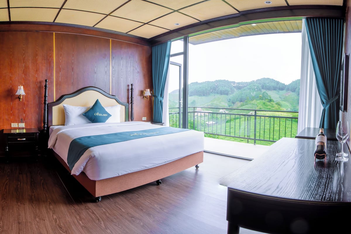 Quarto de hotel com uma cama grande, uma mesa de cabeceira de madeira, uma mesa com garrafa e copo, e uma varanda com vista para colinas verdes.