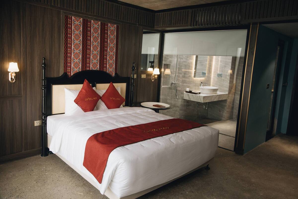 Quarto de hotel com uma cama grande, uma mesa de cabeceira, uma pequena mesa redonda, banheiro com pia e espelho, e uma porta deslizante que dá para outro cômodo.