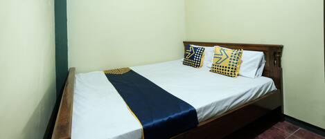 Uma cama de solteiro com estrutura de madeira, roupa de cama branca e almofadas decorativas em um quarto com paredes lisas e chão de azulejos.