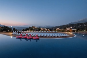 Piscina de resort com espreguiçadeiras e boias de flamingos, cercada por edifícios e montanhas.