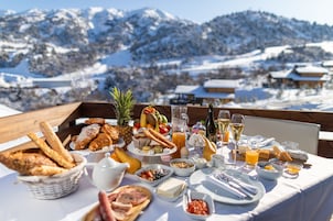 Um buffet de café da manhã com diversos pães, frutas e bebidas em uma mesa com vista para as montanhas ao fundo.