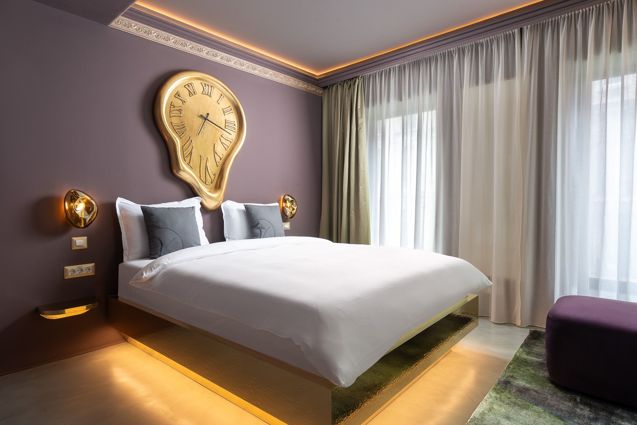 Quarto moderno com cama grande, relógio de parede único e vista para o exterior através de cortinas translúcidas.