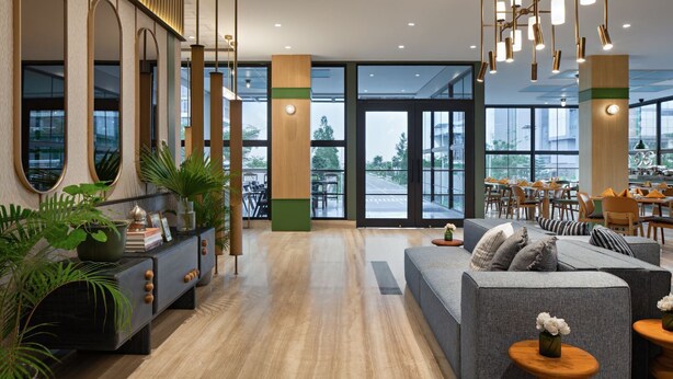 Um lobby moderno com balcão de recepção em madeira, espelhos grandes e uma área de estar com sofás e mesas.