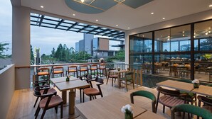 Um restaurante moderno com terraço, mesas e cadeiras de madeira e grandes janelas com vista para a paisagem urbana.