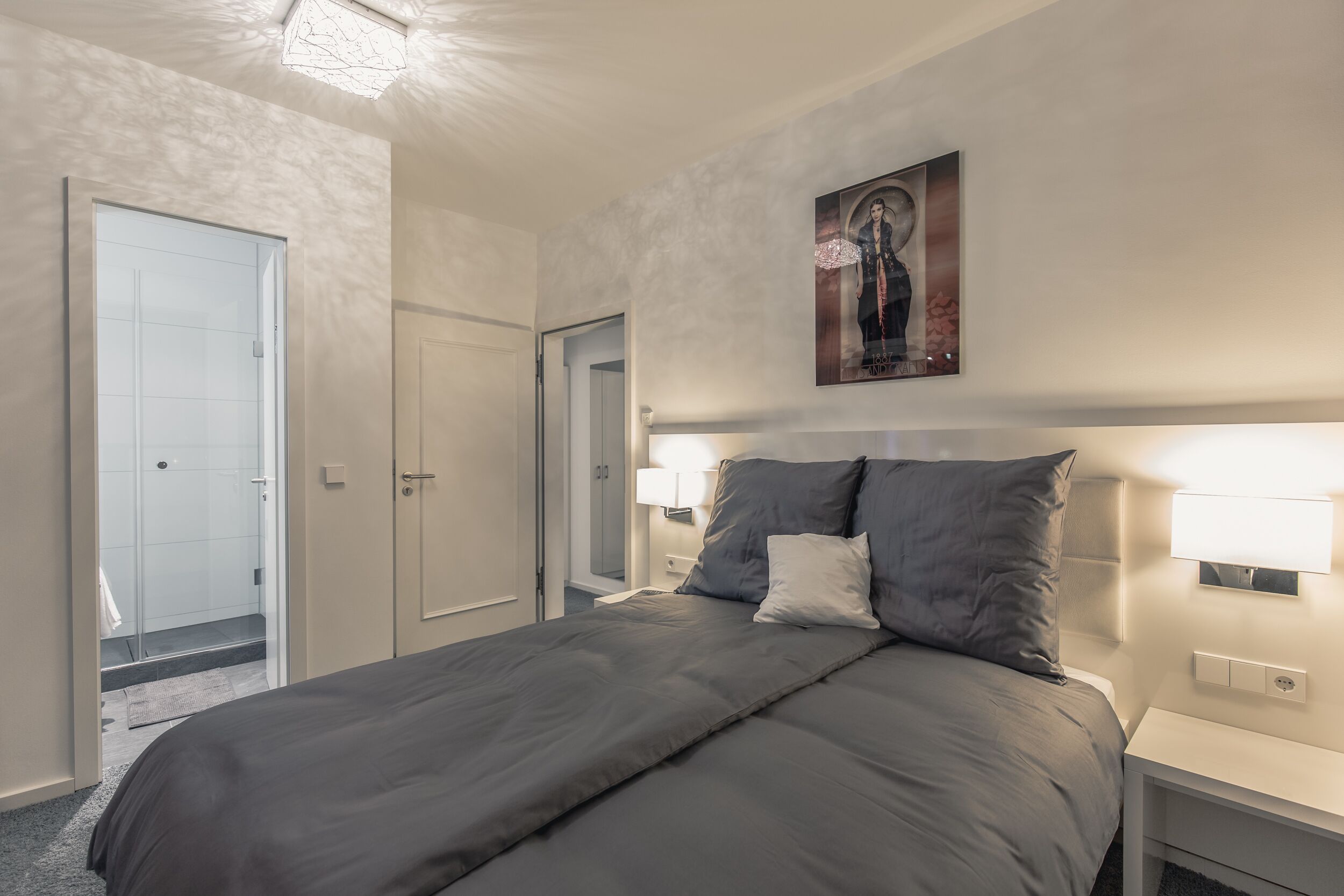 Quarto com cama, abajures, um quadro e porta para o banheiro.