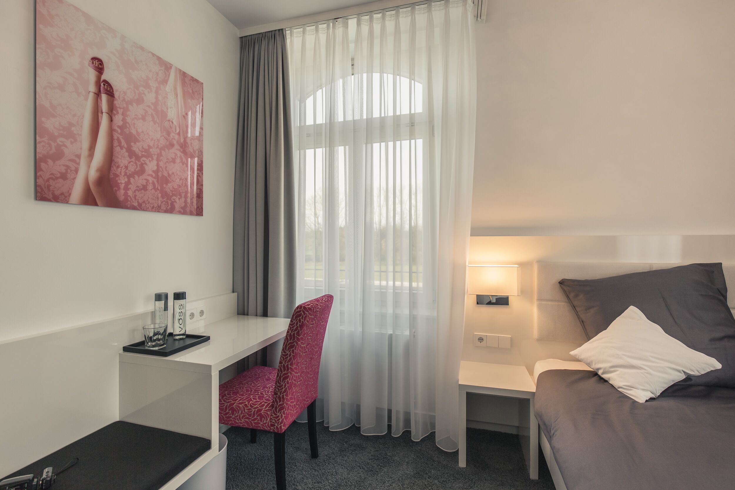 Quarto de hotel com cama, escrivaninha com cadeira, janela grande com cortinas leves e uma obra de arte pendurada na parede.
