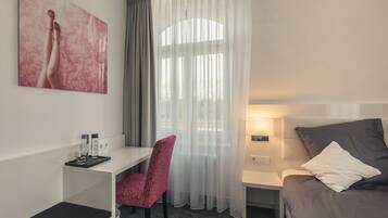 Quarto de hotel com cama, escrivaninha com cadeira, janela grande com cortinas leves e uma obra de arte pendurada na parede.