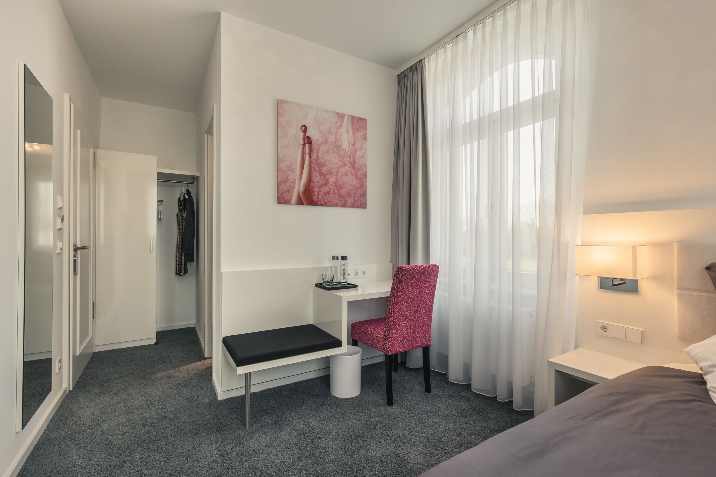 Quarto de hotel com cama, escrivaninha com cadeira, pia pequena e um quadro na parede.
