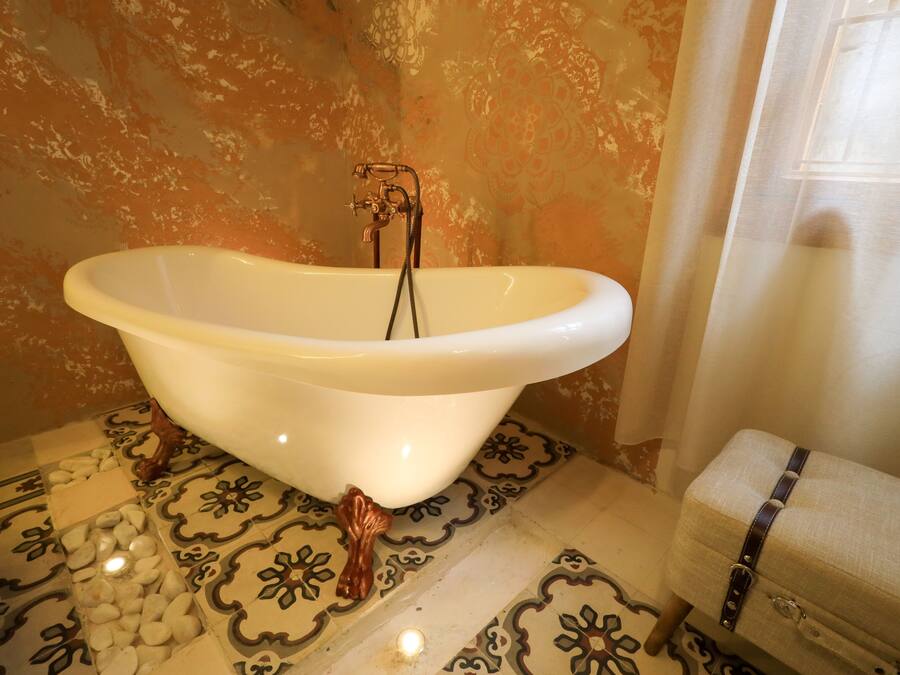 Uma banheira antiga com torneira de latão em um banheiro com azulejos estampados e uma mala vintage.