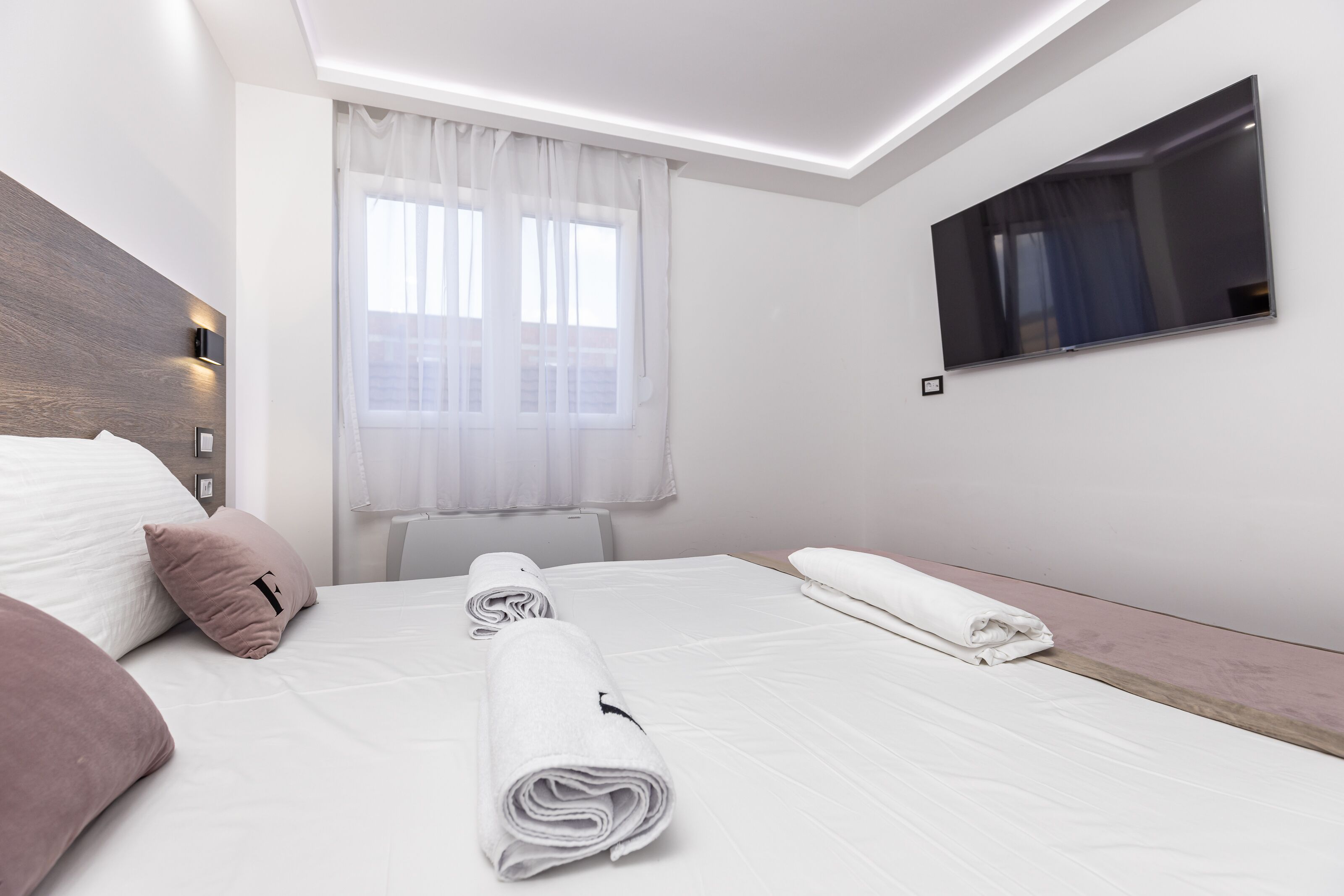 Quarto de hotel moderno com uma cama bem arrumada, uma janela grande com cortinas, uma televisão de tela plana fixada na parede e uma cabeceira de madeira.