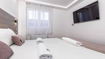 Quarto de hotel moderno com uma cama bem arrumada, uma janela grande com cortinas, uma televisão de tela plana fixada na parede e uma cabeceira de madeira.