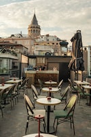 Um café ao ar livre com mesas redondas e cadeiras de vime, uma área de bar e vista para uma torre histórica ao fundo.