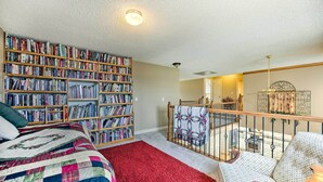 Quarto com uma grande estante de livros, tapete vermelho, cama acolchoada e varanda com uma cadeira.