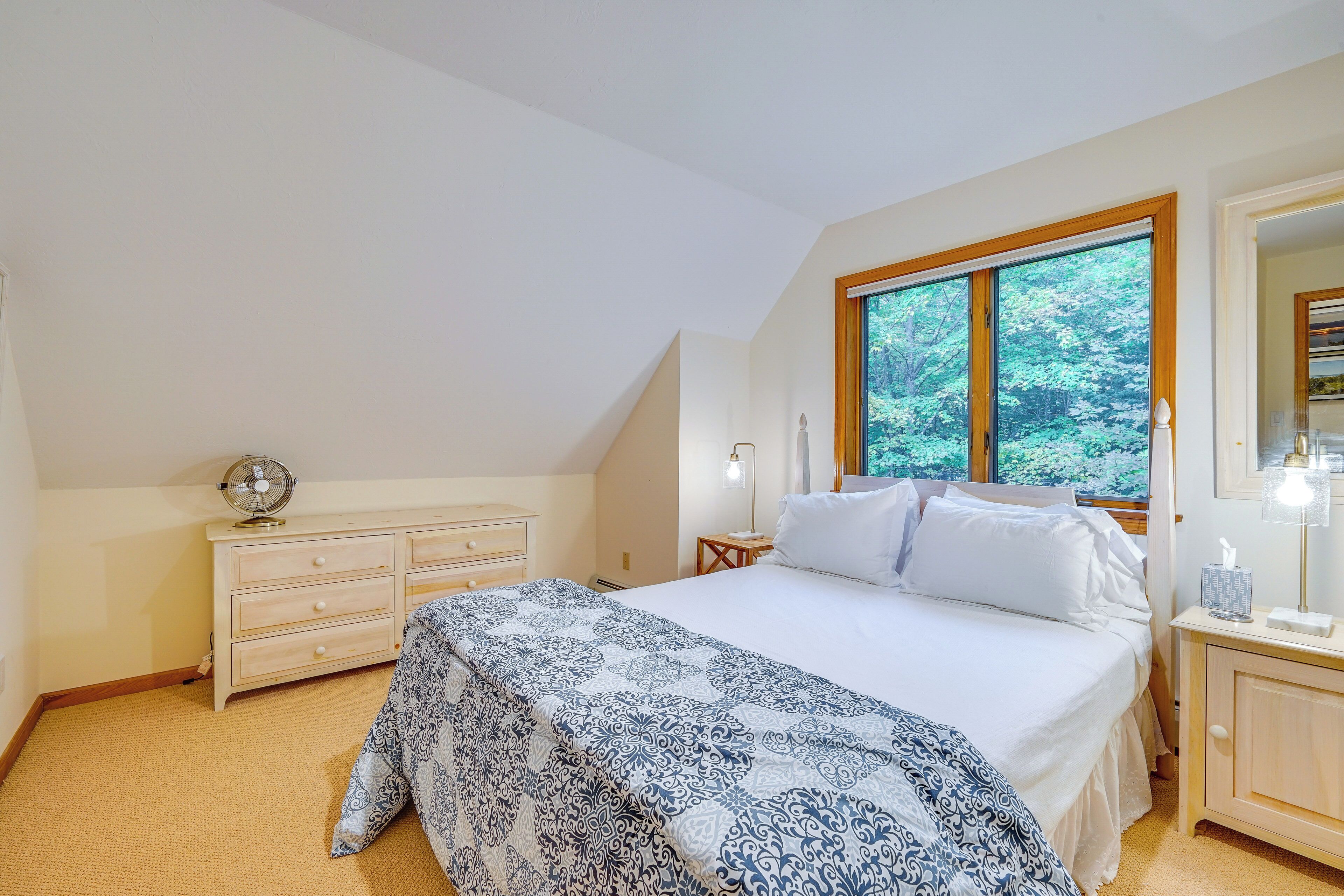 Quarto com cama grande, penteadeira, mesa de cabeceira e janela com vista para árvores.