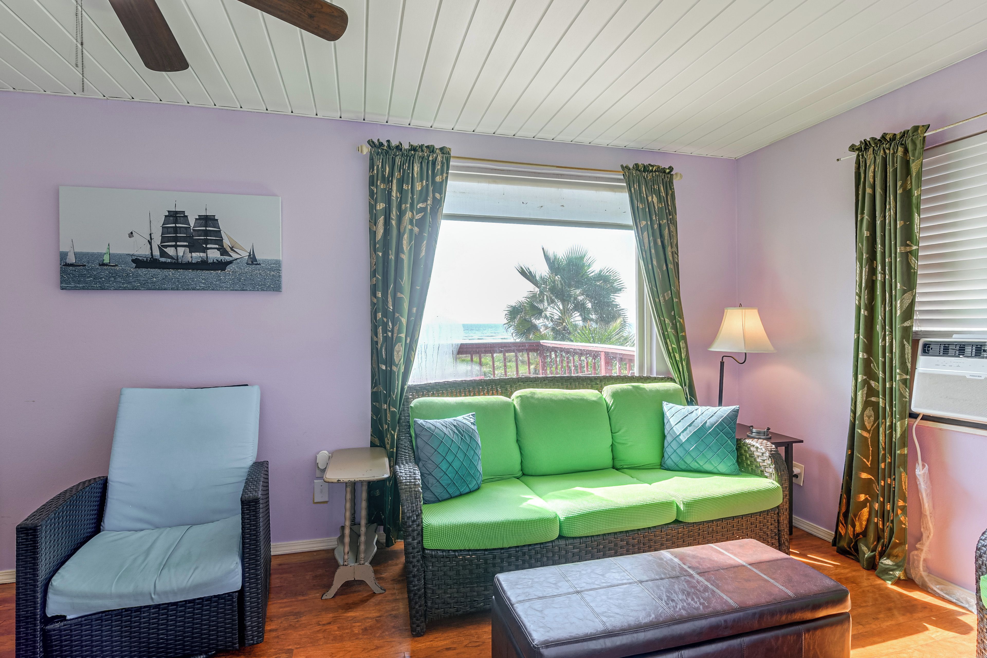 Quarto com sofá verde, cadeira de vime, mesa pequena, abajur e janela com cortinas.