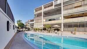 Uma área de piscina com um edifício de vários andares ao fundo, mobiliário de exterior e plantas em vasos.