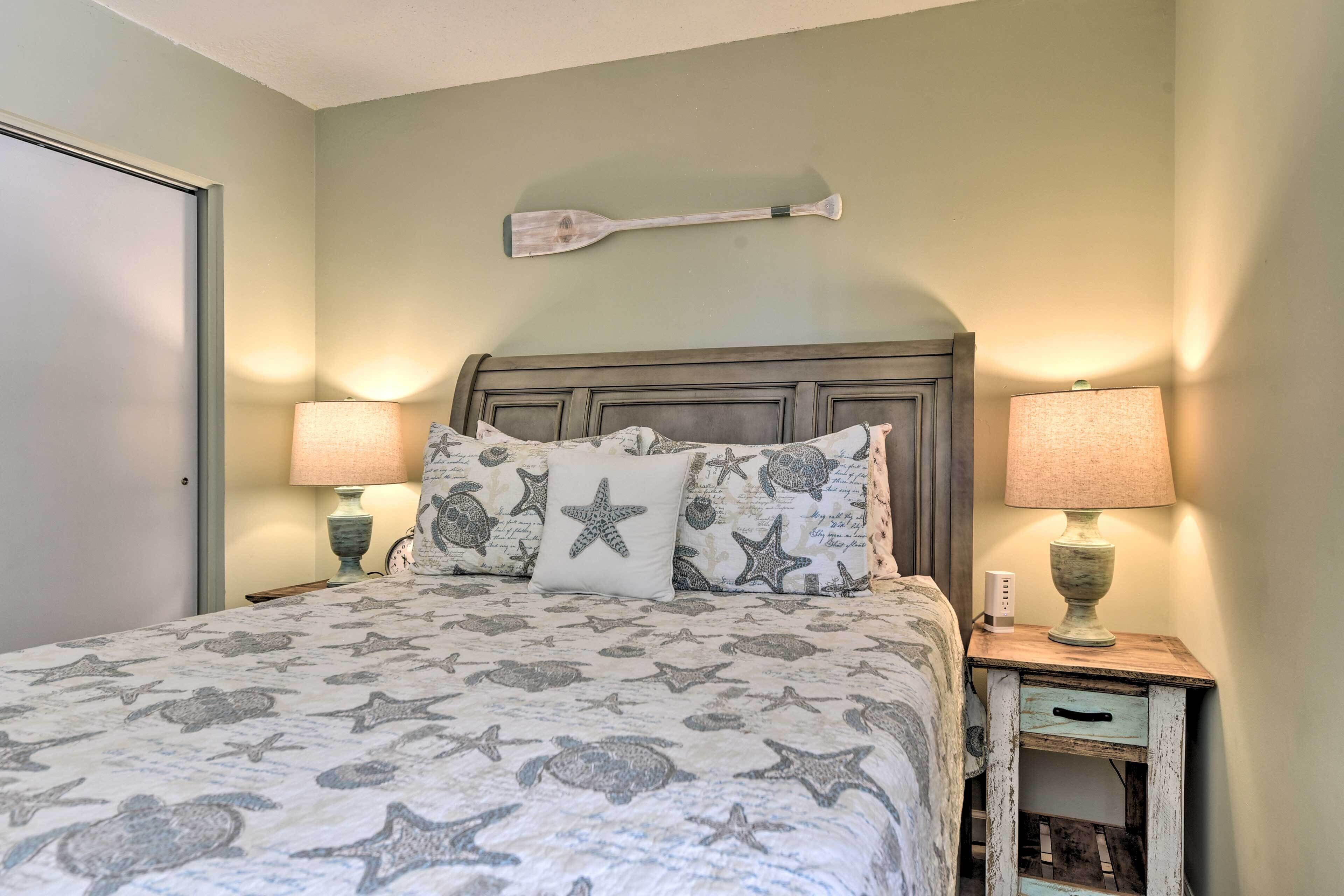 Quarto com cama estampada com padrão de estrela e concha do mar, duas luminárias de cabeceira, mesa de cabeceira de madeira com gaveta e um remo fixado na parede.