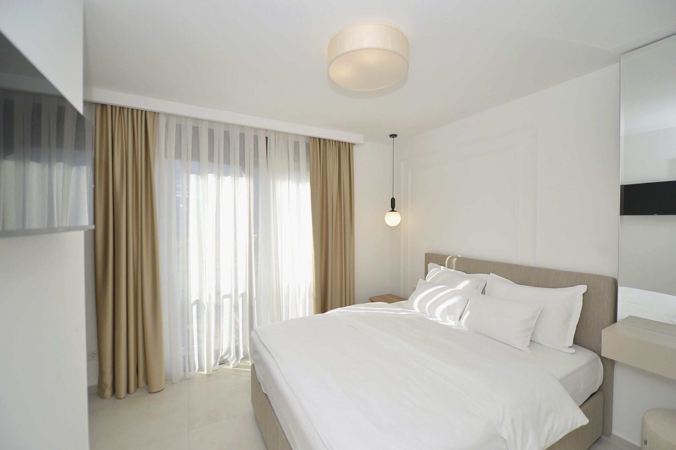 Quarto de hotel moderno com uma cama grande, mesa de cabeceira, luminária pendente e vista para o exterior através de cortinas translúcidas.