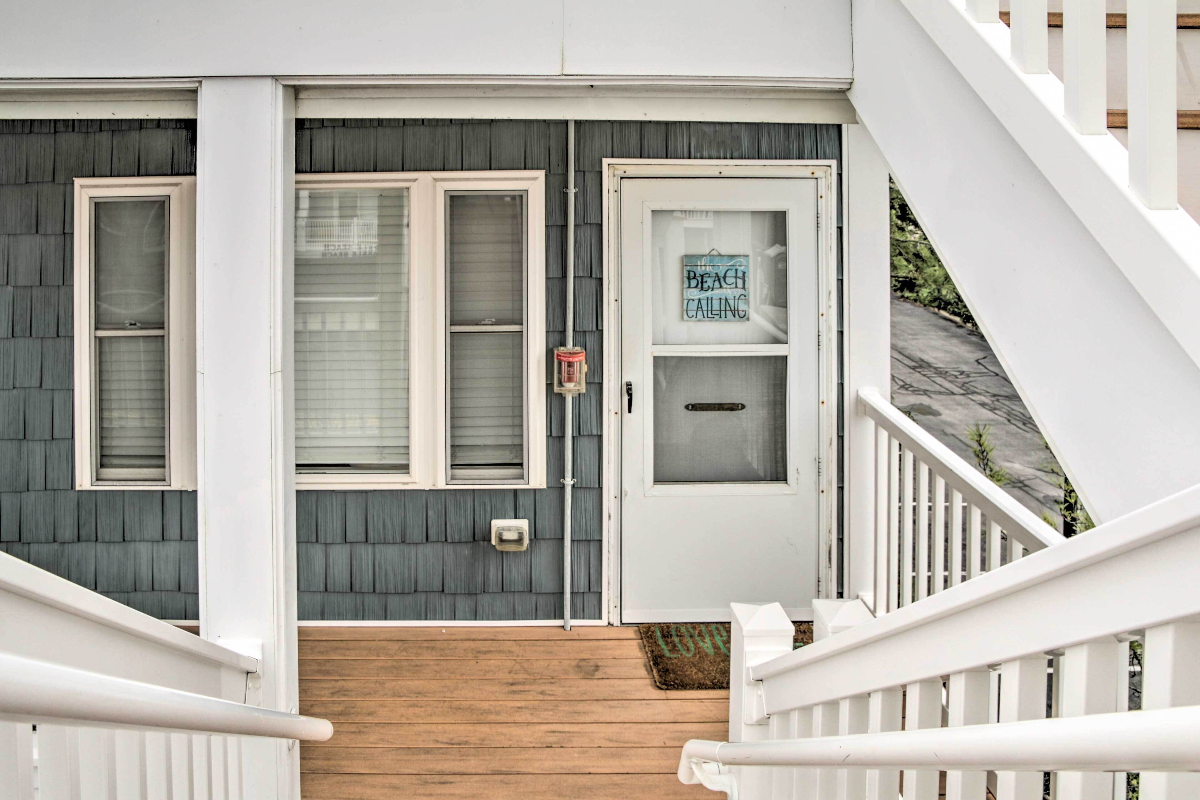 Vista frontal de uma casa com deck de madeira, duas janelas com persianas, uma porta e um letreiro com a inscrição 'Beach Calling', além de um corrimão branco.