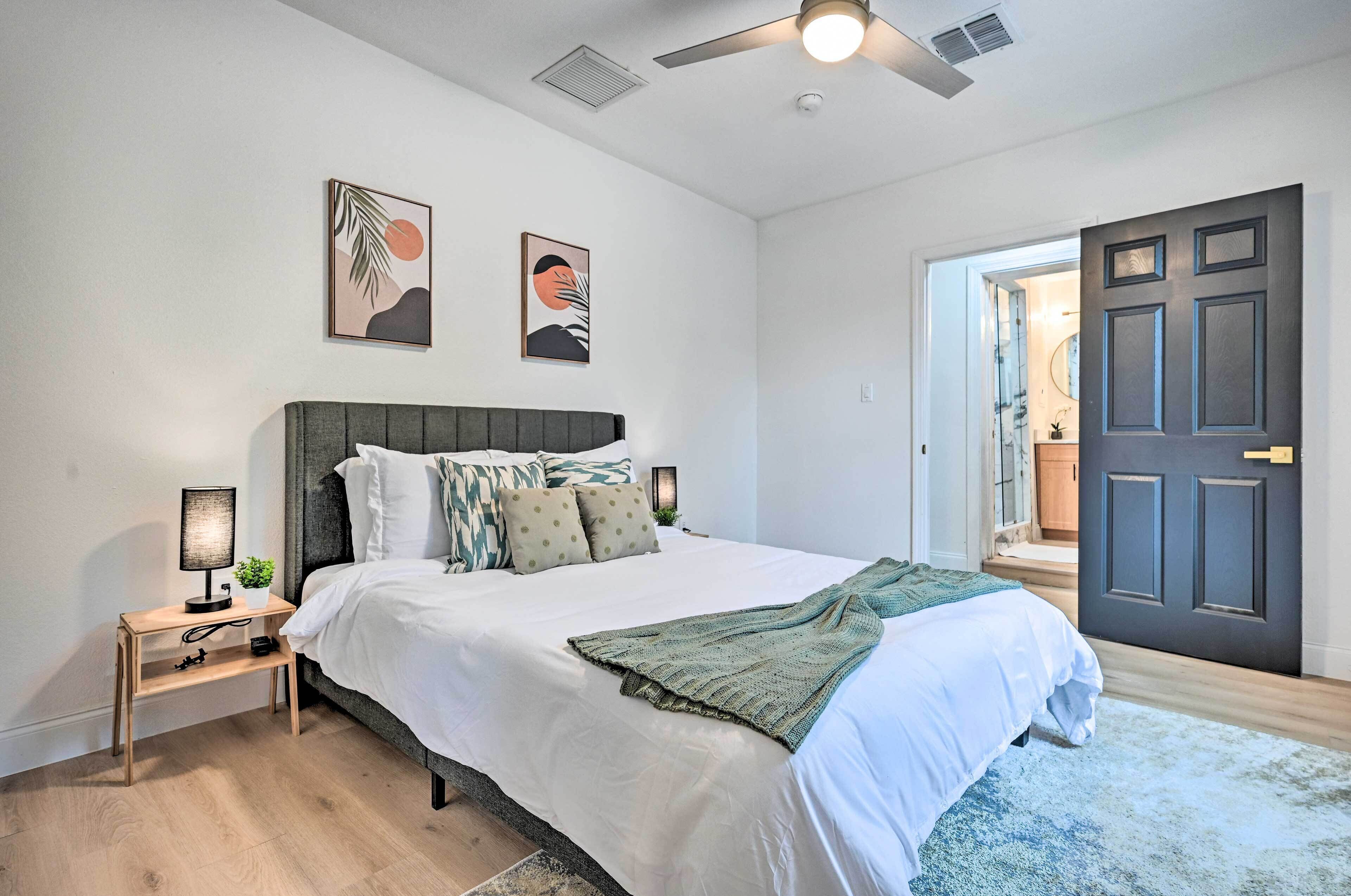 Quarto com cama, mesas de cabeceira, um criado-mudo, ventilador de teto e uma porta.