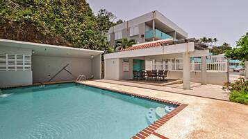 Piscina com deck revestido, área de estar coberta com cadeiras e um edifício com varanda.