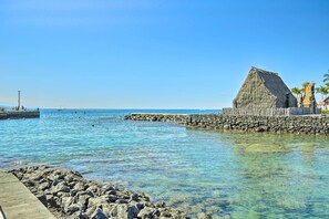 Mar azul e límpido com um edifício de telhado de palha e uma parede de pedra.