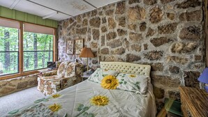 Quarto com parede de pedra, cama com roupa de cama floral, uma cadeira, um abajur e vista para árvores pela janela.