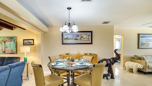 Uma sala de jantar com uma mesa redonda, cadeiras e um candelabro. Há um sofá e uma mesa de centro ao primeiro plano, e um quadro na parede.