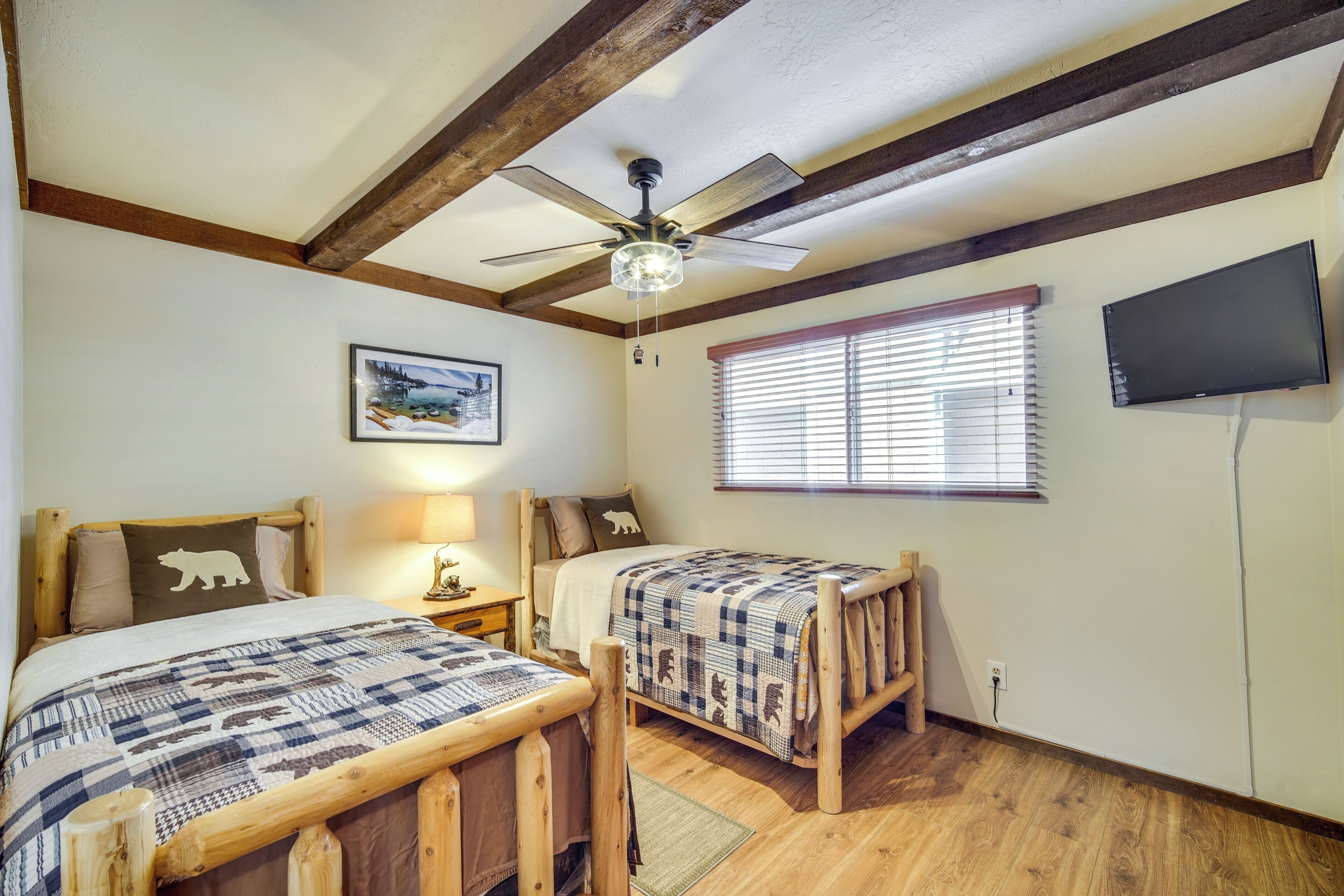 Quarto com duas camas de madeira, ventilador de teto, televisão montada e janela com persianas.