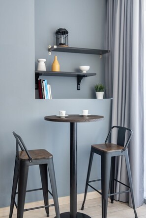 Quarto moderno com parede cinza, uma mesa redonda de madeira e duas cadeiras pretas de metal, uma estante de metal preta com objetos decorativos, e uma cortina cinza.