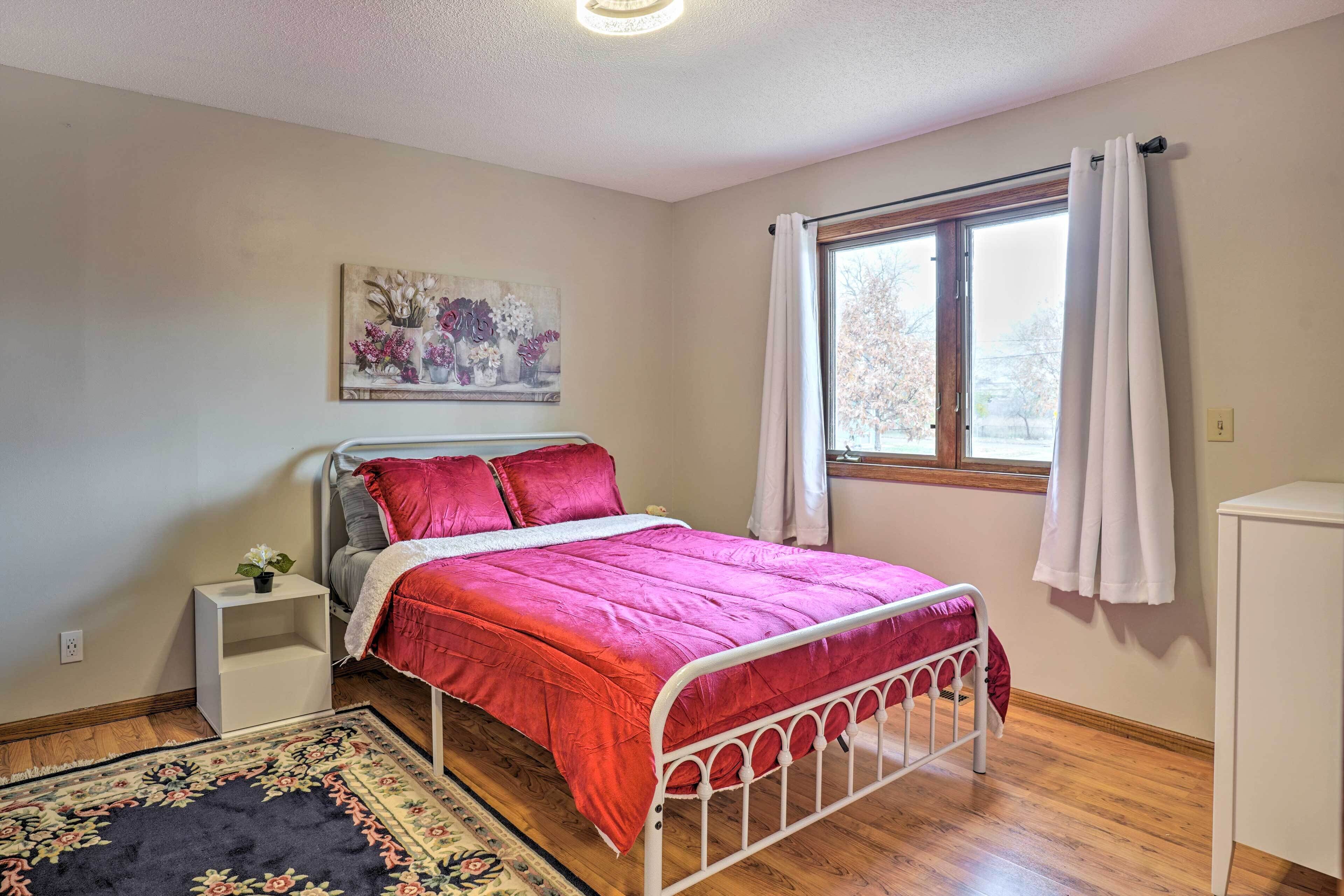 Um quarto com cama, mesa de cabeceira, janela com cortinas, um quadro na parede e um armário pequeno.