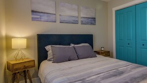 Quarto com cama, mesa de cabeceira com abajur, gaveta de madeira e porta azul.