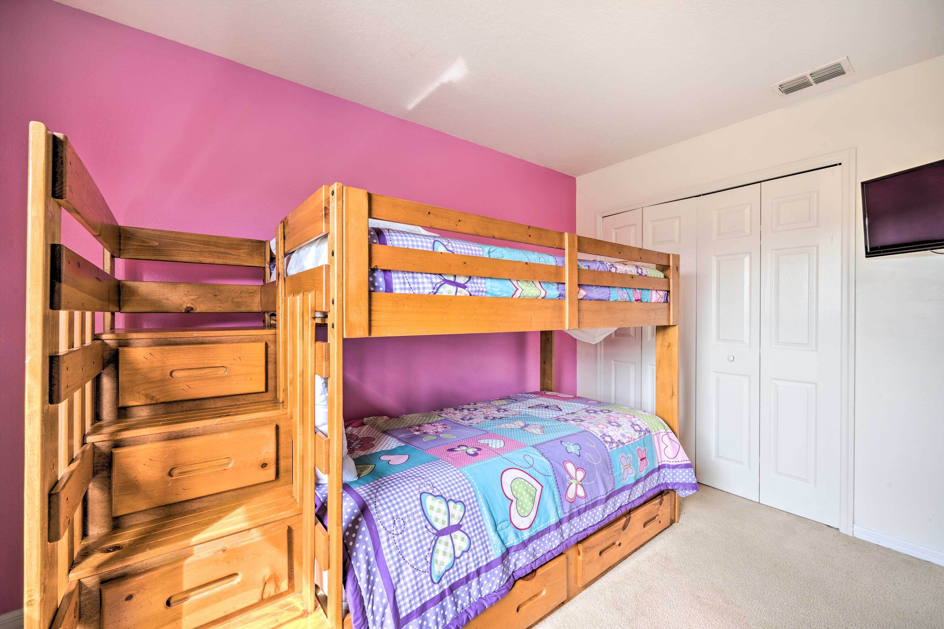 Uma cama de solteiro com edredom roxo e azul, gavetas de madeira e uma porta branca ao fundo.