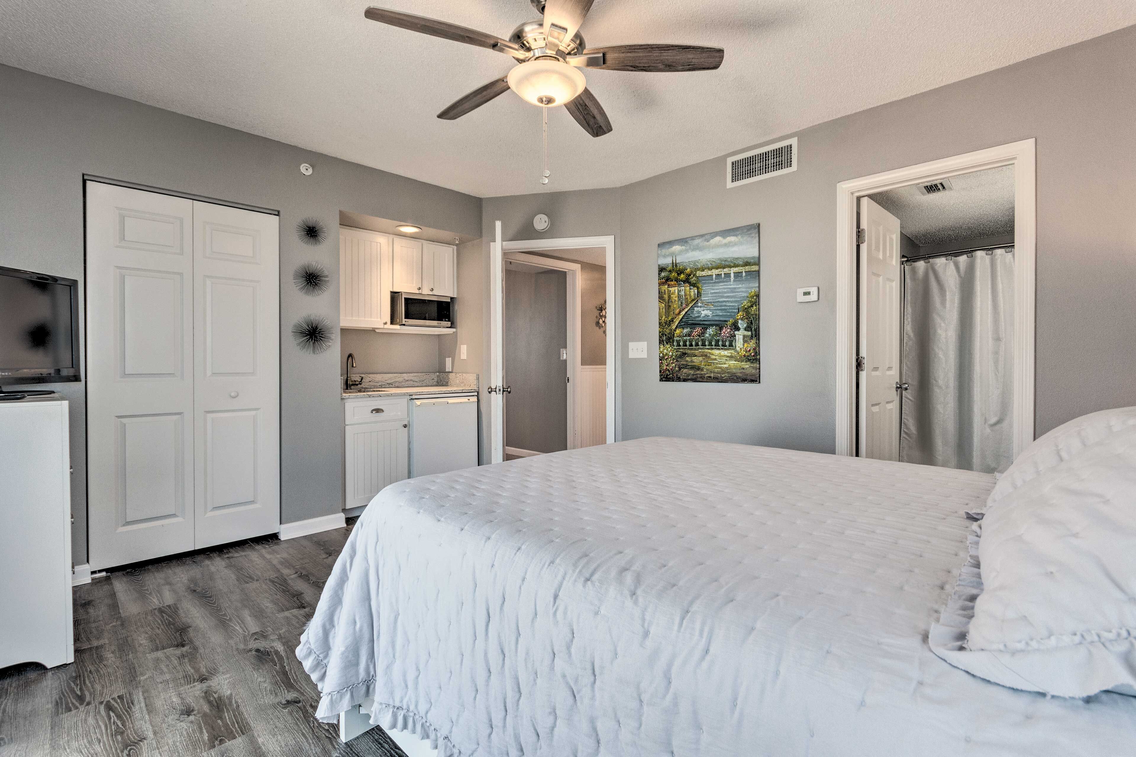Quarto com cama grande, ventilador de teto, cozinha compacta e porta para outro cômodo.