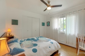 Um quarto com cama, ventoinha no teto, janela com cortinas e uma cadeira.