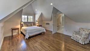 Quarto com teto inclinado, cama com colcha branca, piso de madeira, mesa de cabeceira, abajur, cadeira e porta que dá para um banheiro.