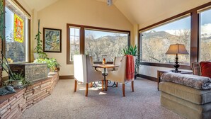 Quarto aconchegante com mesa de jantar, cadeiras, sofá e grandes janelas com vista para as montanhas.