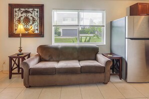 Uma sala de estar com sofá, mesa, abajur, janela com persianas e geladeira.