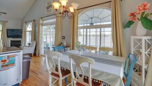 Uma sala de jantar com uma mesa posta para quatro pessoas, rodeada por cadeiras brancas, um lustre e grandes janelas com cortinas.