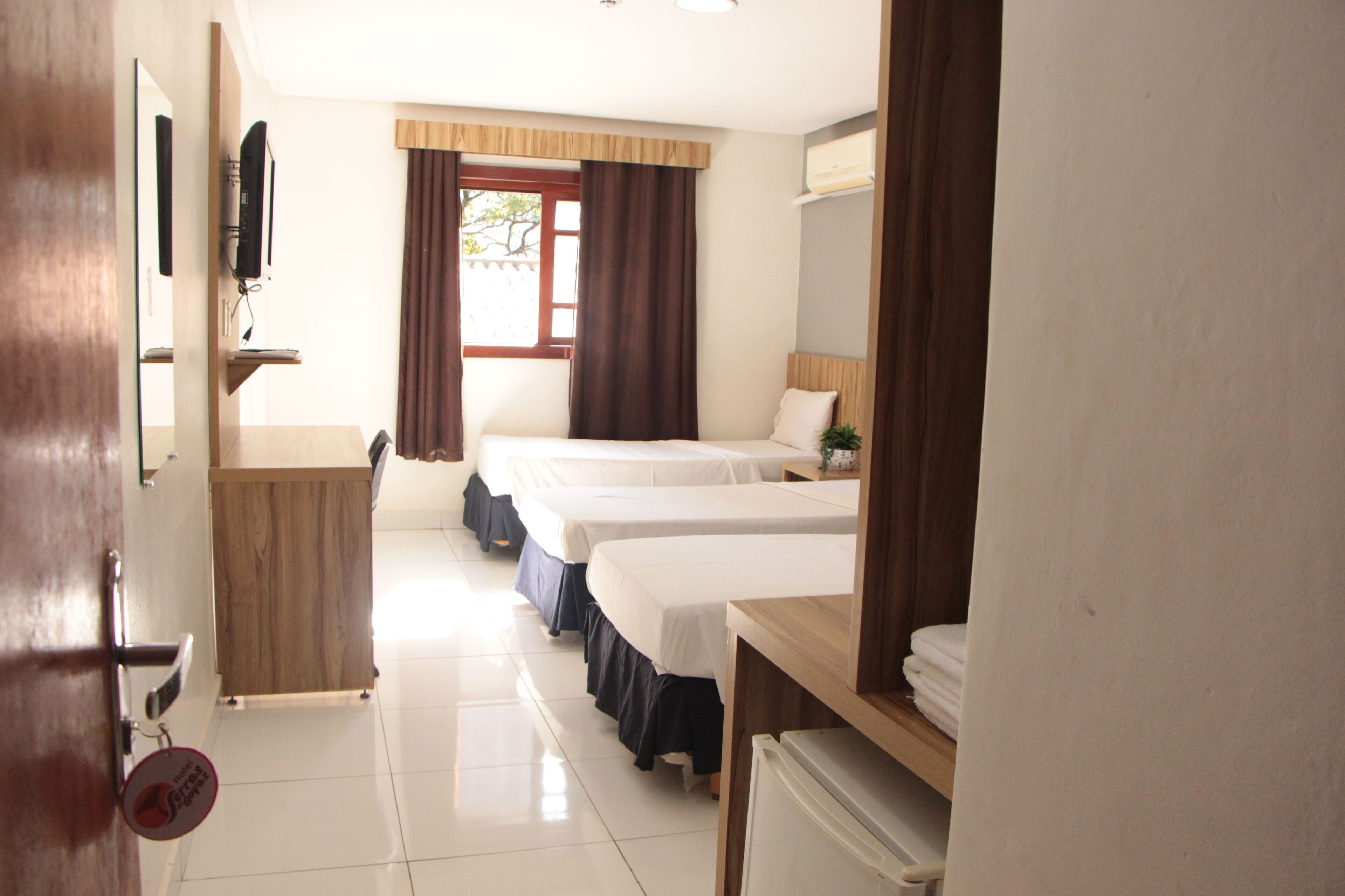 Quarto de hotel com três camas, uma escrivaninha de madeira, televisão e uma janela com cortinas.