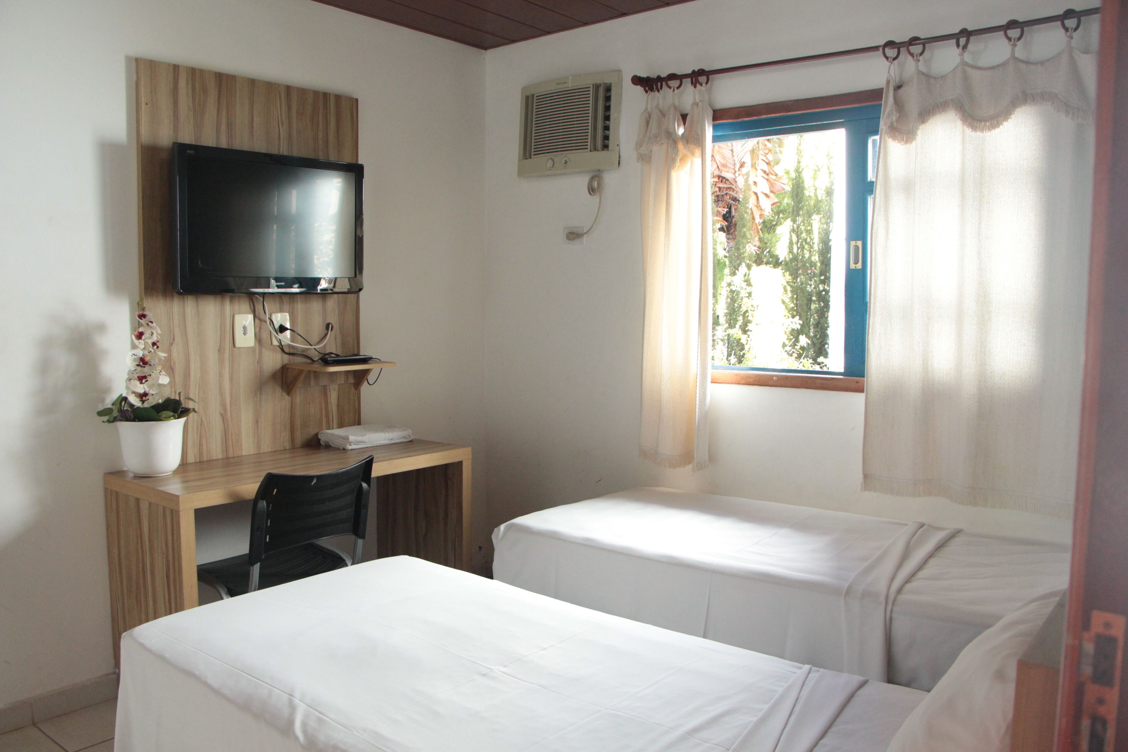 Quarto com duas camas, uma escrivaninha com cadeira, televisão fixada na parede e janela com cortinas.