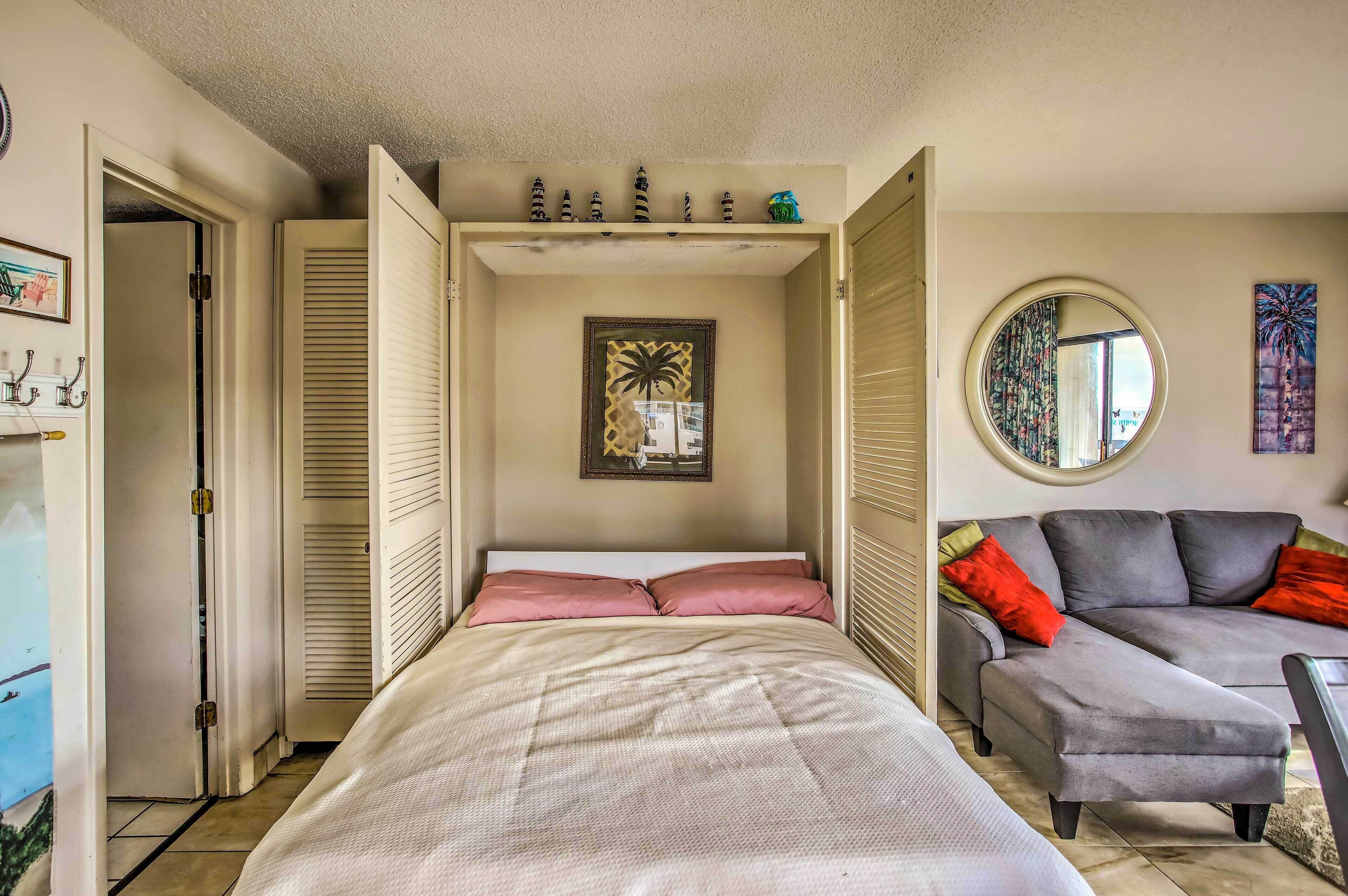 Um quarto com uma cama, um sofá cinza, um espelho redondo e um quadro na parede.