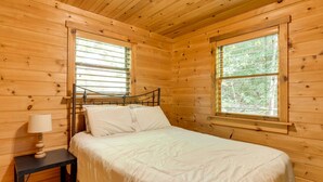 Quarto de cabana de madeira com uma cama, uma mesinha de cabeceira e duas janelas.