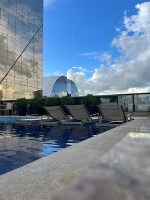 Uma piscina com espreguiçadeiras, um edifício moderno com fachada de vidro e uma estrutura em forma de cúpula ao fundo.