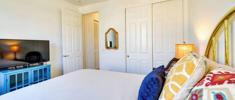 Quarto com cama, televisão sobre um móvel azul, ventoinha no teto e portas duplas brancas.