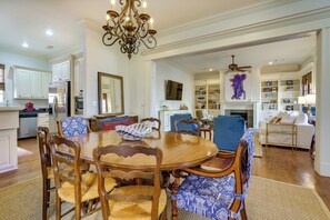 Uma ampla sala de jantar com mesa e cadeiras de madeira, um candelabro e vista para a cozinha e sala de estar.
